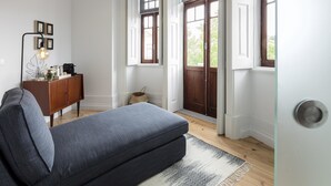 Um quarto amplo com teto alto, um sofá cinza escuro, um armário de madeira e uma escrivaninha com abajur.
