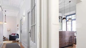 Um corredor com piso de madeira, paredes brancas e tapete cinza, conduzindo a um espaço com um banco de madeira e uma janela.