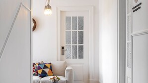 Um saguão de hotel moderno com decoração em tons de branco, um sofá com almofadas coloridas, uma mesa de centro de madeira e um lustre decorativo.