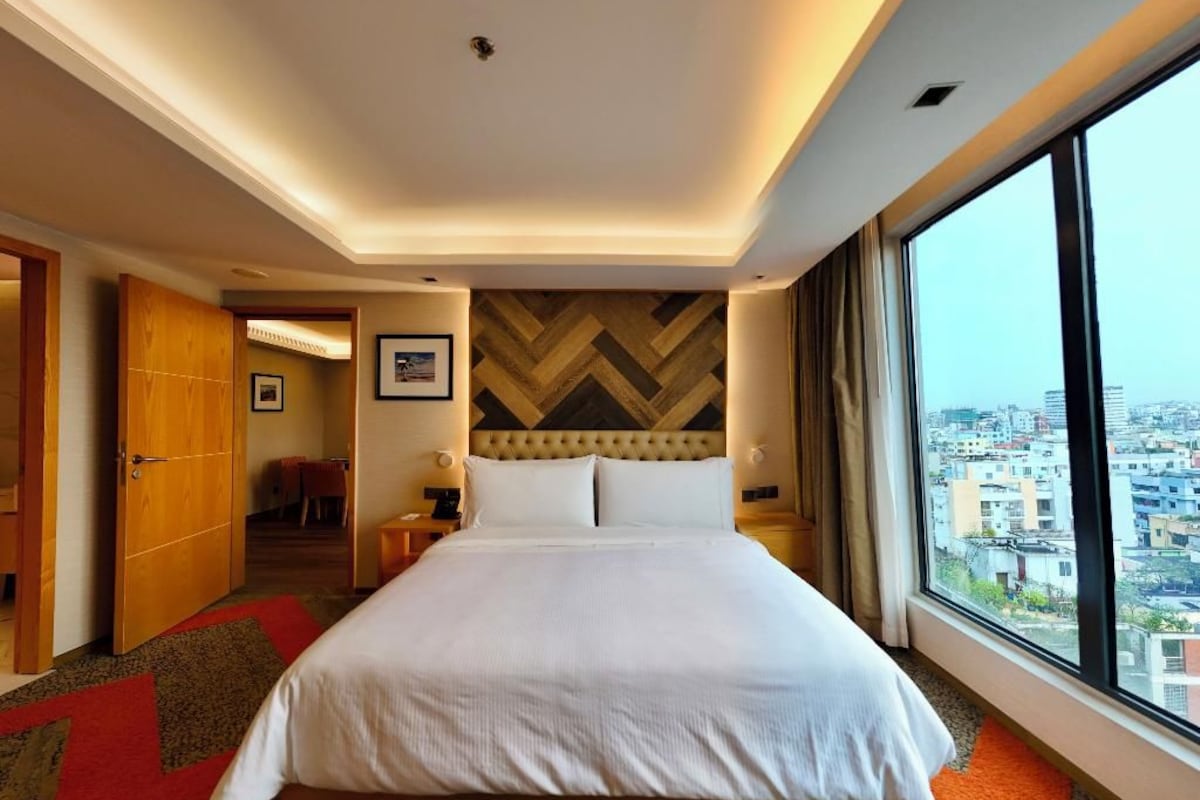 Quarto de hotel com uma cama grande, vista da cidade pela janela e parede com padrão losango.