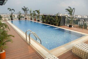 Uma piscina infinita no terraço com vista para a cidade, rodeada por plantas em vasos e um deck de madeira.