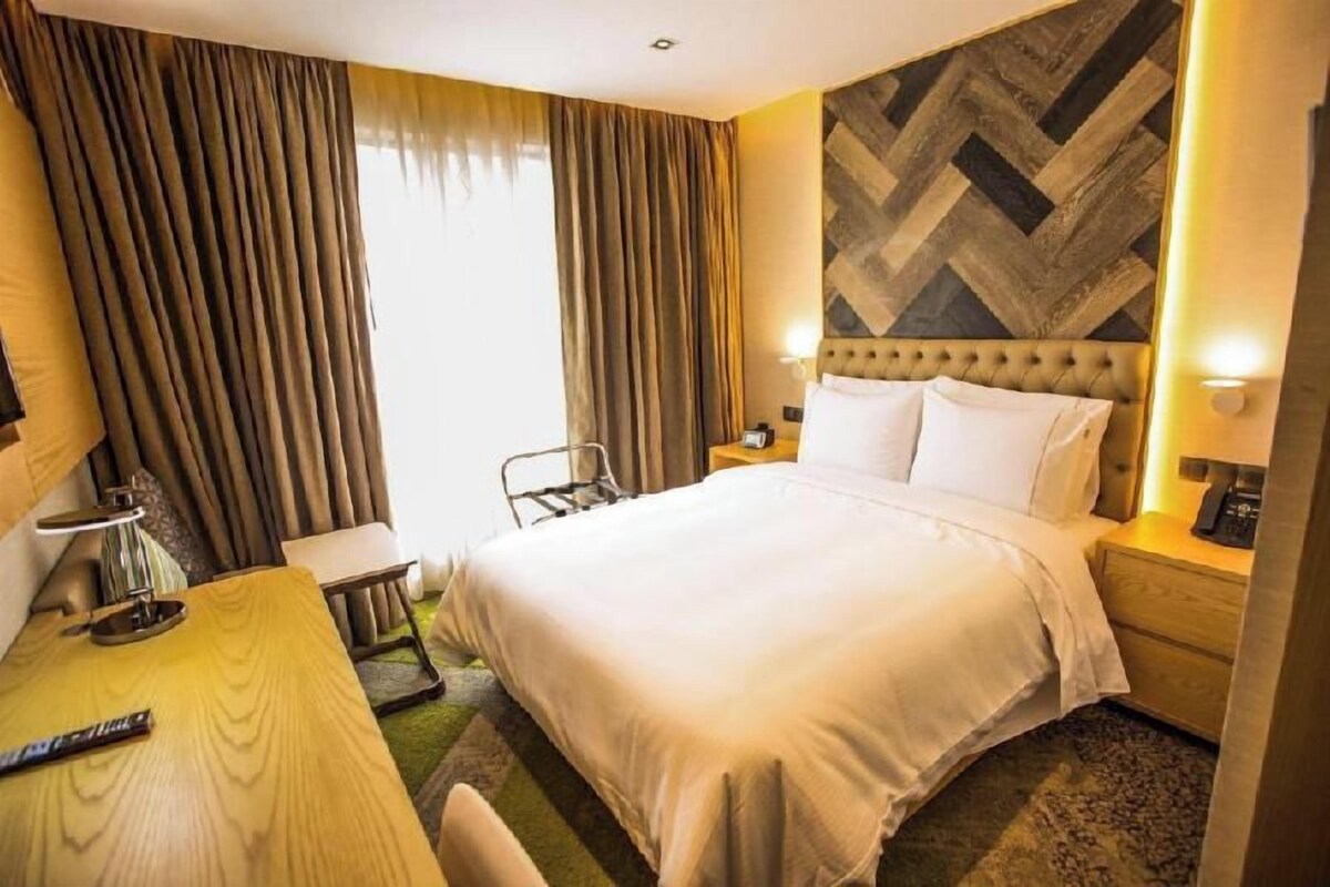 Quarto de hotel com cama, mesas de cabeceira, escrivaninha, cadeira e cortinas.