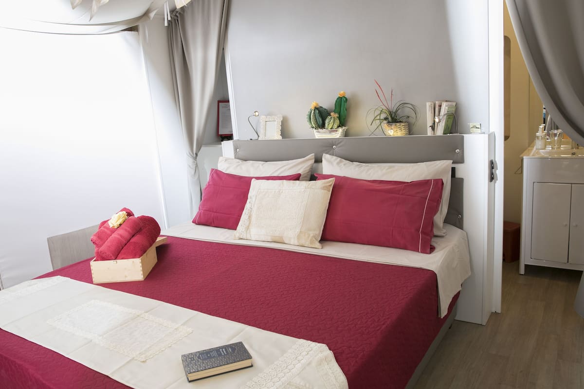 Cama bem arrumada com colcha vermelha e branca, mesa de cabeceira de madeira com abajur e uma pequena planta no criado-mudo.