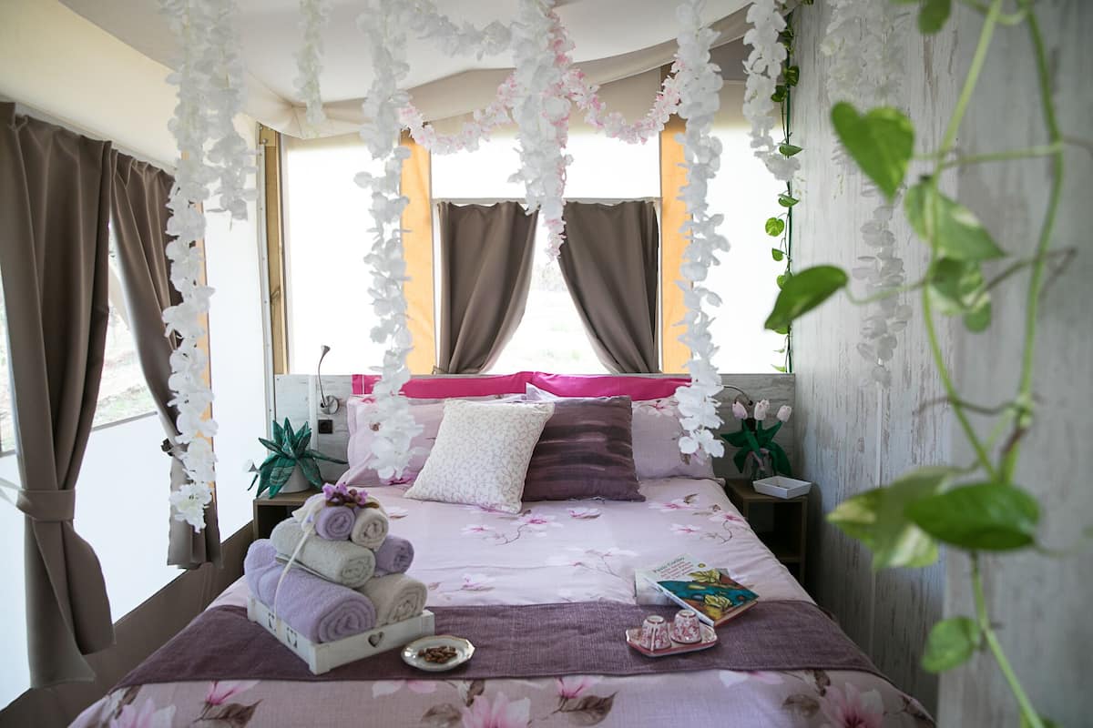 Quarto com cama, almofadas, mesa de cabeceira, janela com cortinas e uma planta.