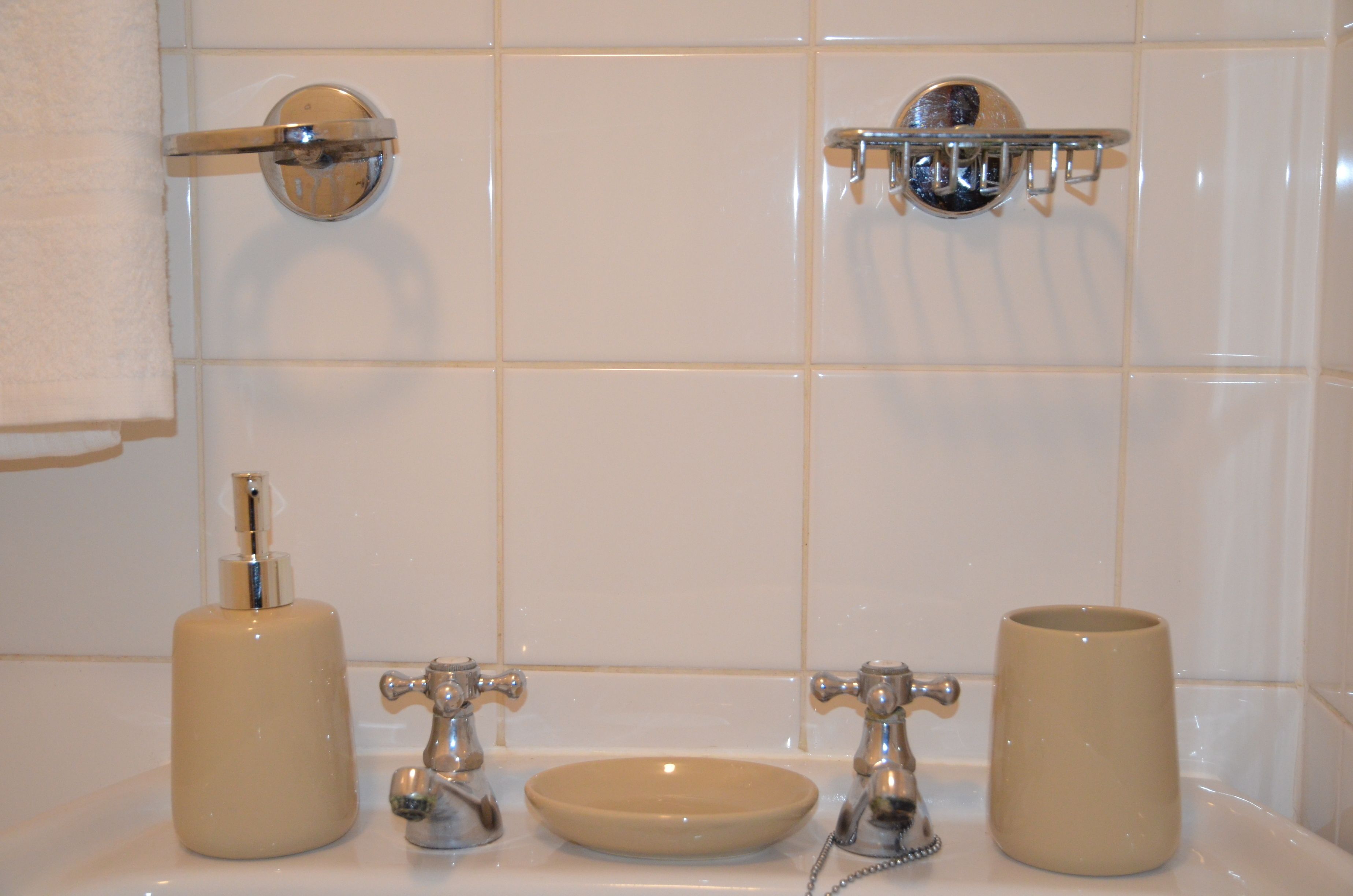 Casa de banho com azulejos brancos, uma pia com duas torneiras, dispensador de sabonete, uma chávena e um porta-toalhas.
