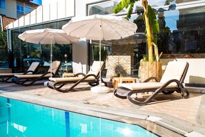 Área de piscina com espreguiçadeiras e guarda-sóis, parede de tijolos e um edifício de vidro ao fundo.