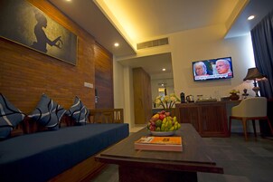 Uma sala de estar moderna com um sofá, uma mesa de centro com um livro e uma fruteira, uma TV exibindo notícias e um armário de madeira.