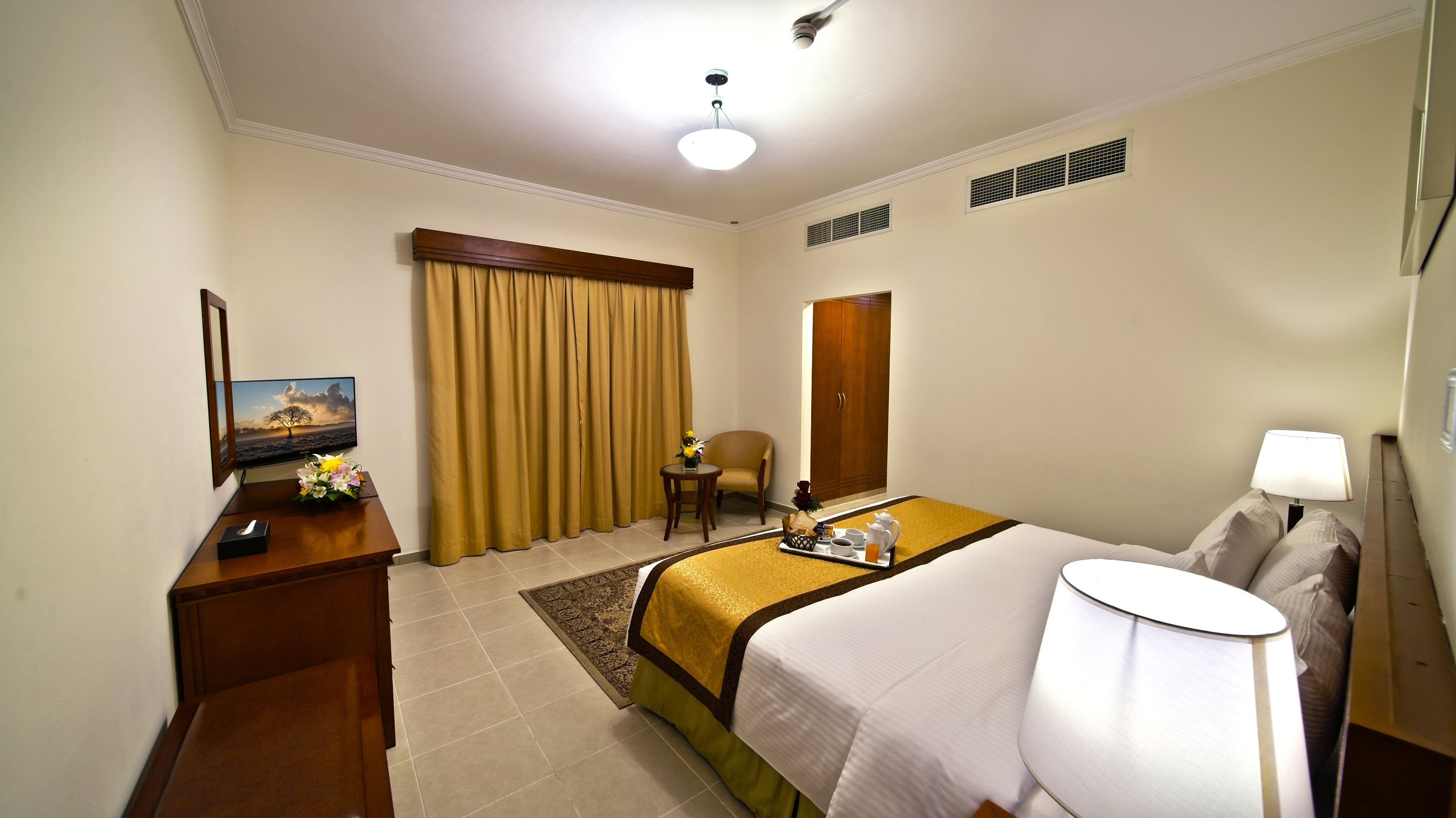 Quarto de hotel com cama, escrivaninha com abajur, cadeira, mesa pequena com um vaso e janela com cortinas.