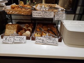 Vitrine de padaria com diversos produtos de panificação, como pão, croissants e muffins, cada um com seu nome e ingredientes indicados.