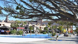 Uma área de piscina pública com pessoas nadando e relaxando, cercada por edifícios e palmeiras.