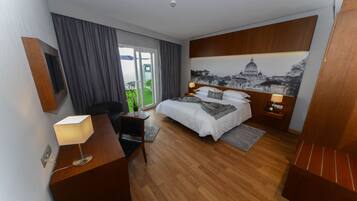 Quarto de hotel com uma cama grande, uma escrivaninha com luminária, uma cadeira, uma pequena mesa com uma planta e vista para a cidade por uma porta deslizante.
