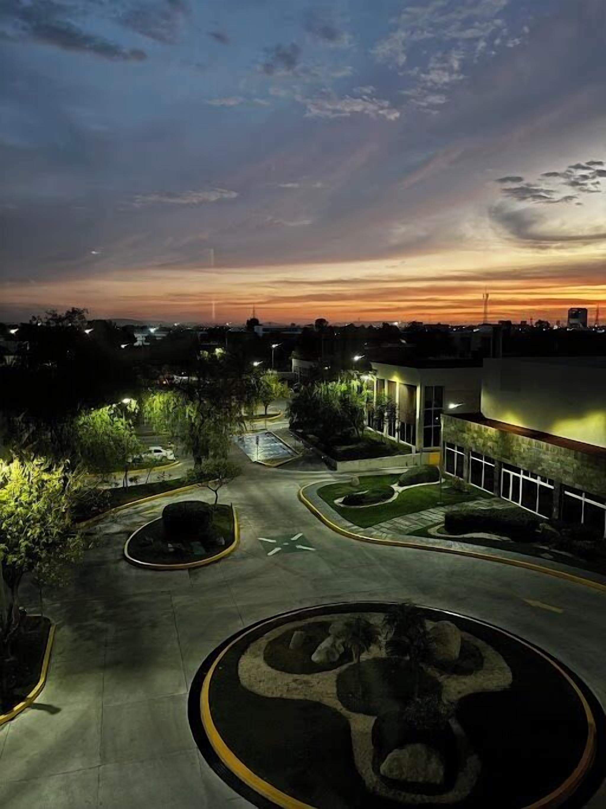 Uma área de estacionamento ampla com um design circular central, rodeada por vegetação e edifícios modernos.