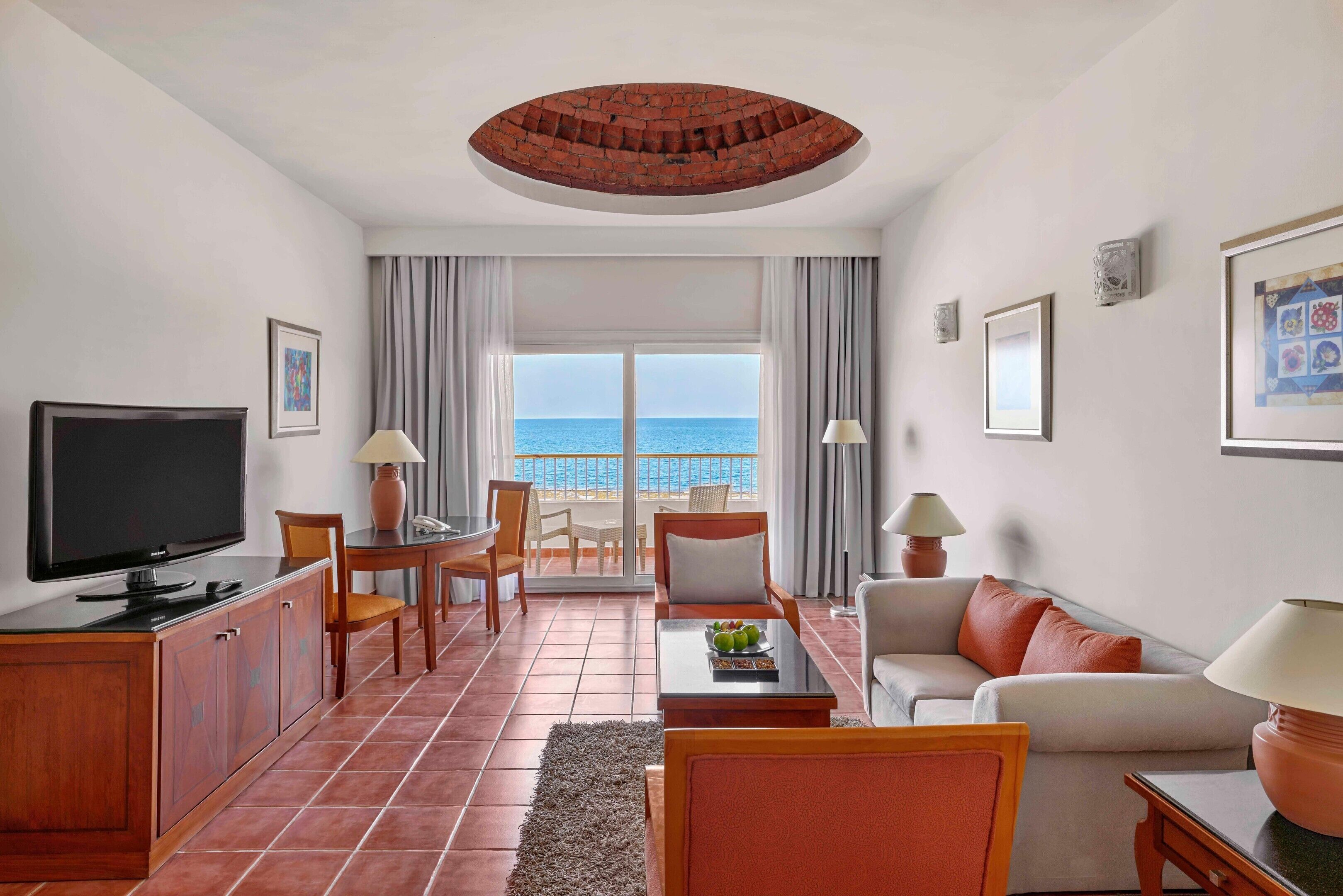 Quarto de hotel com televisão, área de estar e varanda com vista para o mar.