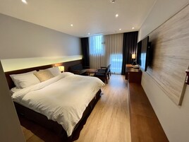 Quarto de hotel com uma cama grande, uma escrivaninha com cadeira, uma televisão e uma janela com cortinas.