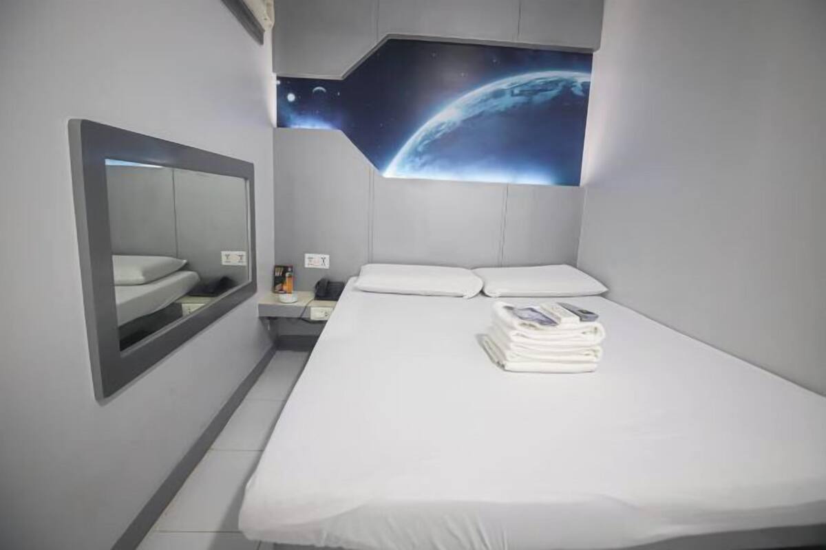 Quarto de hotel moderno com uma cama grande, uma mesa de cabeceira, um espelho e uma TV embutida na parede exibindo uma imagem com tema espacial.