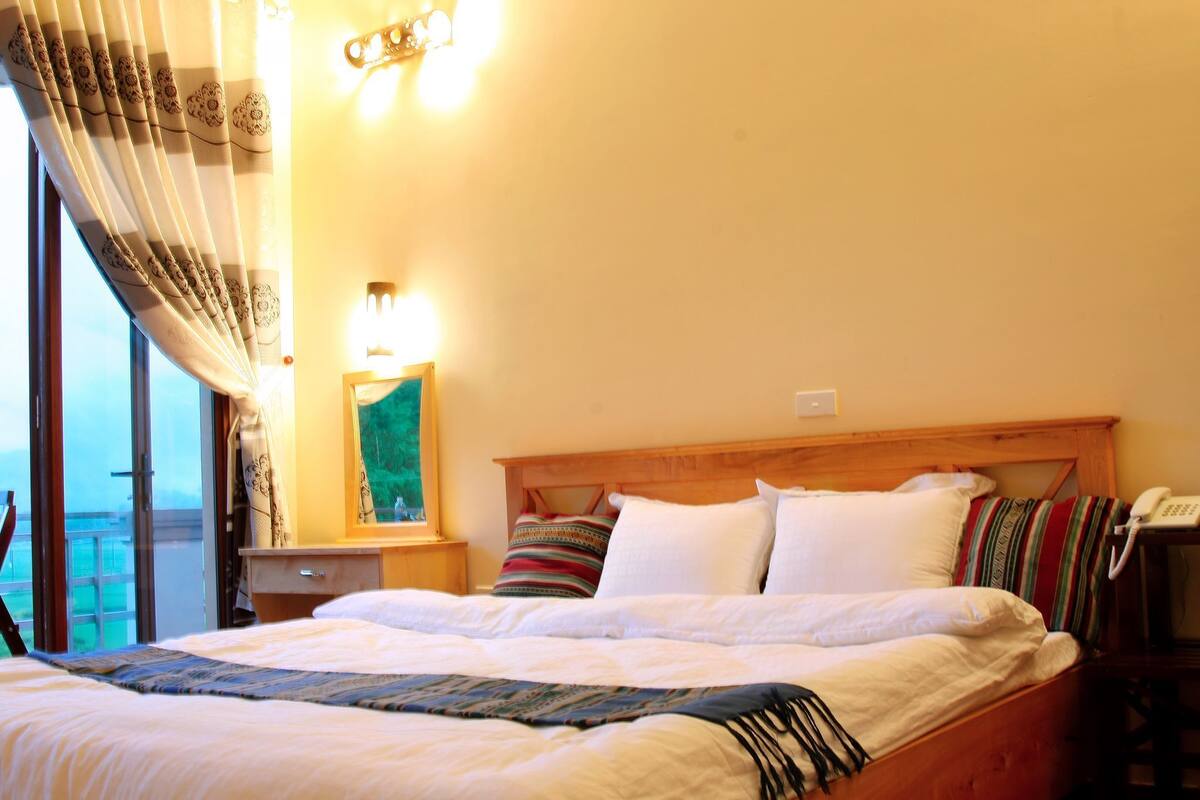 Quarto de hotel com cama, mesa de cabeceira, abajur e vista para o exterior.