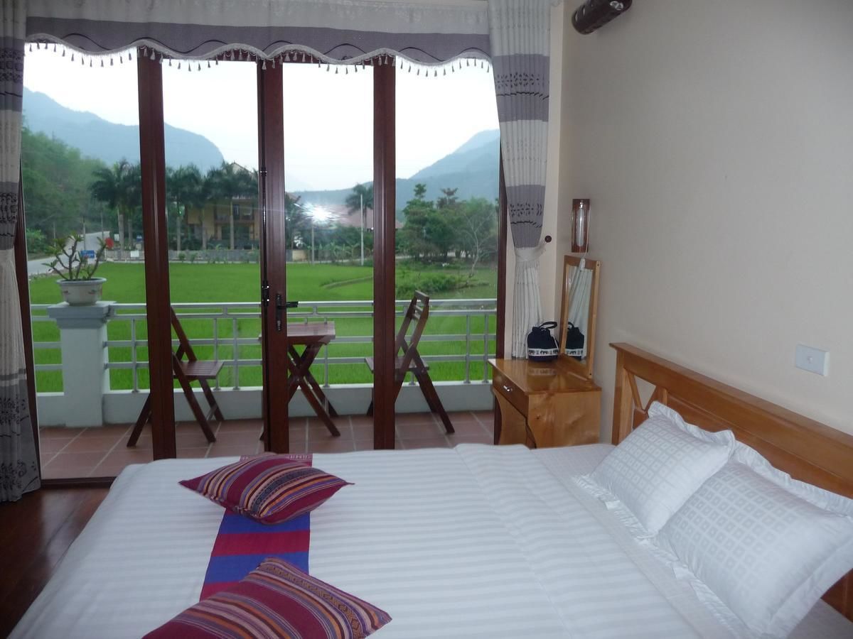 Quarto de hotel com uma cama grande, uma mesa de cabeceira de madeira, uma cadeira e vista para uma paisagem verde através de amplas janelas.