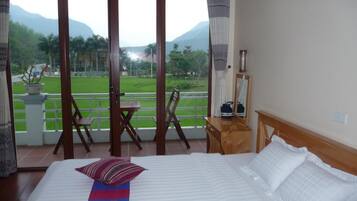 Quarto de hotel com uma cama grande, uma mesa de cabeceira de madeira, uma cadeira e vista para uma paisagem verde através de amplas janelas.