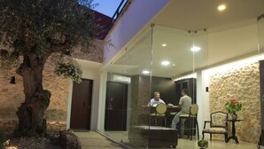 Uma área de refeições moderna ao ar livre, com estrutura de vidro, parede de pedra e uma grande árvore.
