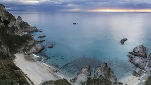 Paisagem costeira com falésias rochosas, águas azul-claras e uma praia arenosa.