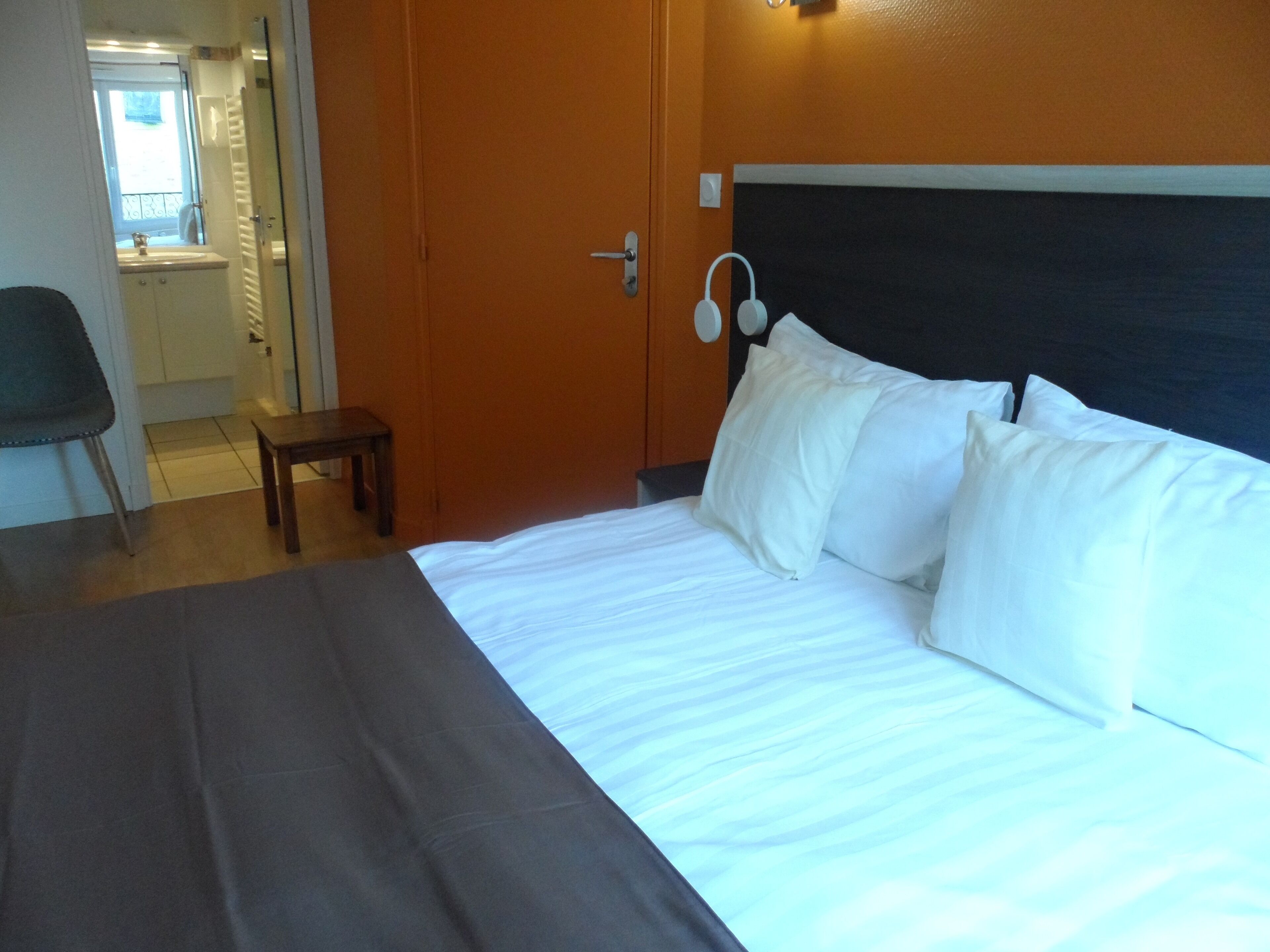 Quarto de hotel com cama, mesa de cabeceira, cadeira e banheiro visível através de uma porta aberta.