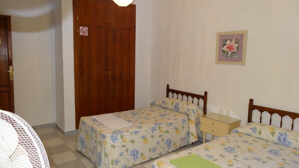 Um quarto com duas camas de solteiro, um guarda-roupa de madeira, uma pequena mesa de cabeceira, um quadro pendurado na parede e um ventilador de teto.