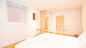 Quarto de hotel moderno com uma cama grande, uma janela com persianas, um espelho e um banheiro visível através de uma porta aberta.