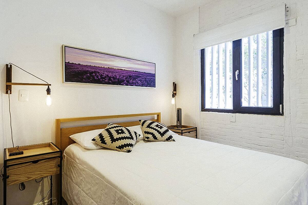Quarto com cama de madeira, duas almofadas estampadas, mesa de cabeceira com abajur, quadro na parede e janela com persianas.