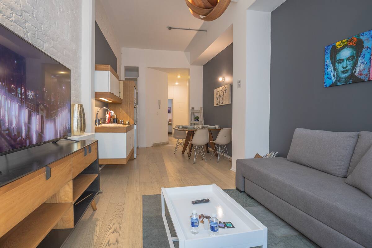 Uma sala de estar moderna com uma televisão de tela plana, um sofá cinza e uma mesa de centro com controles remotos e garrafas de água.