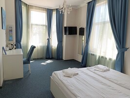 Quarto de hotel com cama, escrivaninha, cadeira, televisão e lustre.