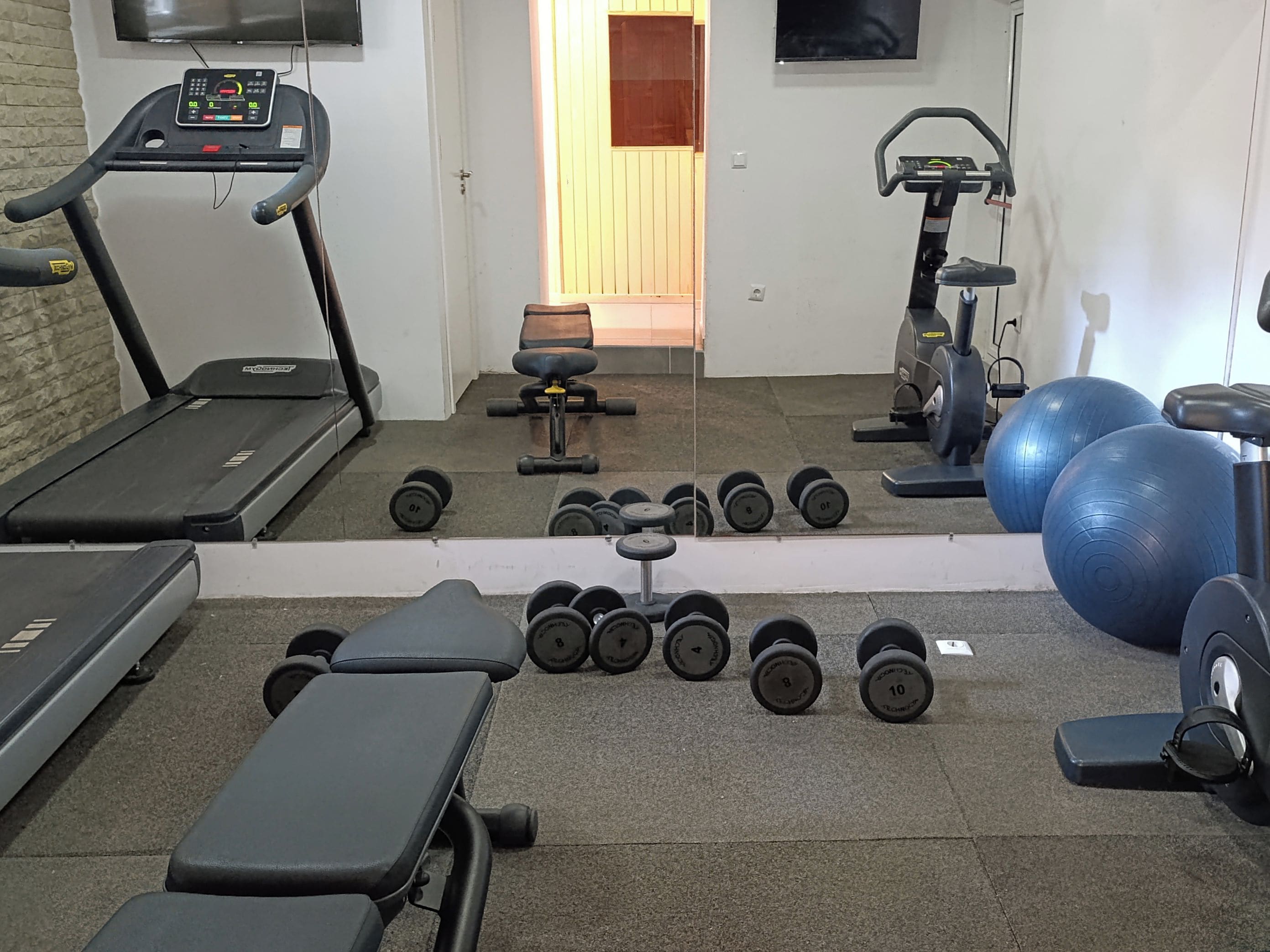 Academia com esteiras, halteres, um banco e bolas de exercício.