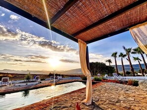 Área da piscina com espreguiçadeiras, um pérgola e vista para palmeiras e montanhas.