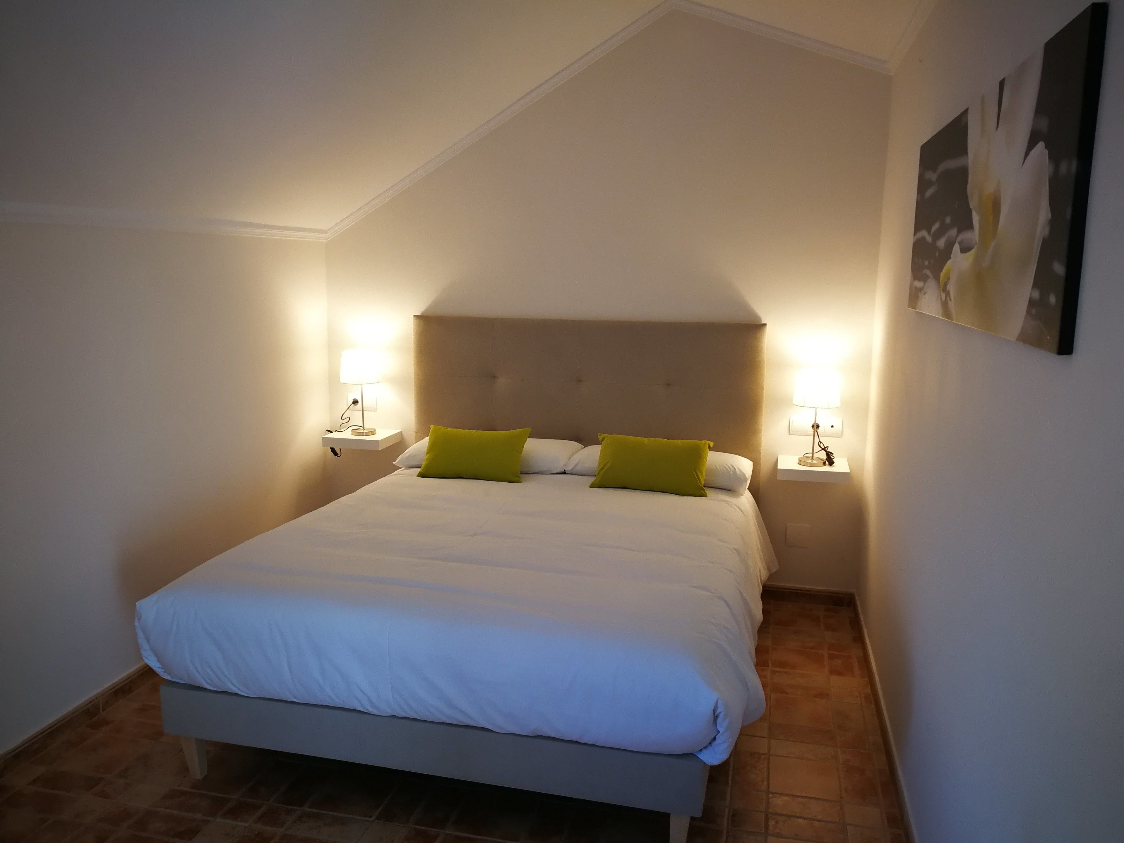 Uma cama bem arrumada com roupa de cama branca e duas almofadas verdes, com duas luminárias de cabeceira em mesas de cabeceira brancas.