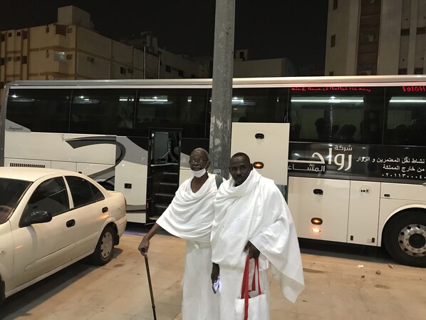 Duas pessoas em frente a um ônibus com texto em árabe na lateral, um carro branco estacionado por perto e um edifício ao fundo.