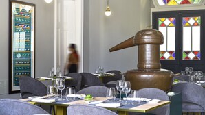 Uma sala de jantar com mesas preparadas para o serviço, um alambique de cobre grande e uma janela de vitrais.