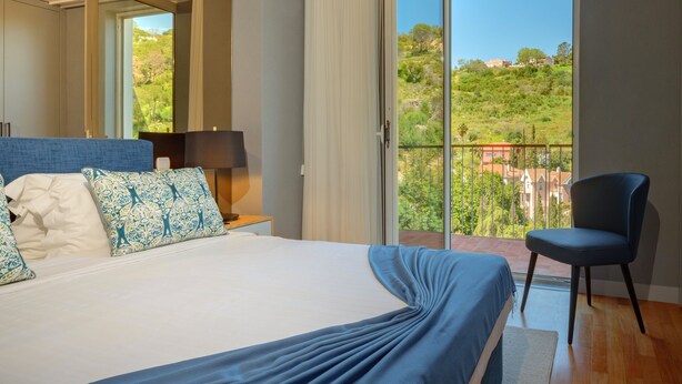 Quarto com cama, poltrona, mesa de cabeceira, luminária e varanda com vista para vegetação e edifícios.