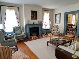 Uma sala de estar tradicional com lareira, dois poltronas, um sofá e uma área de jantar visível por uma porta aberta.