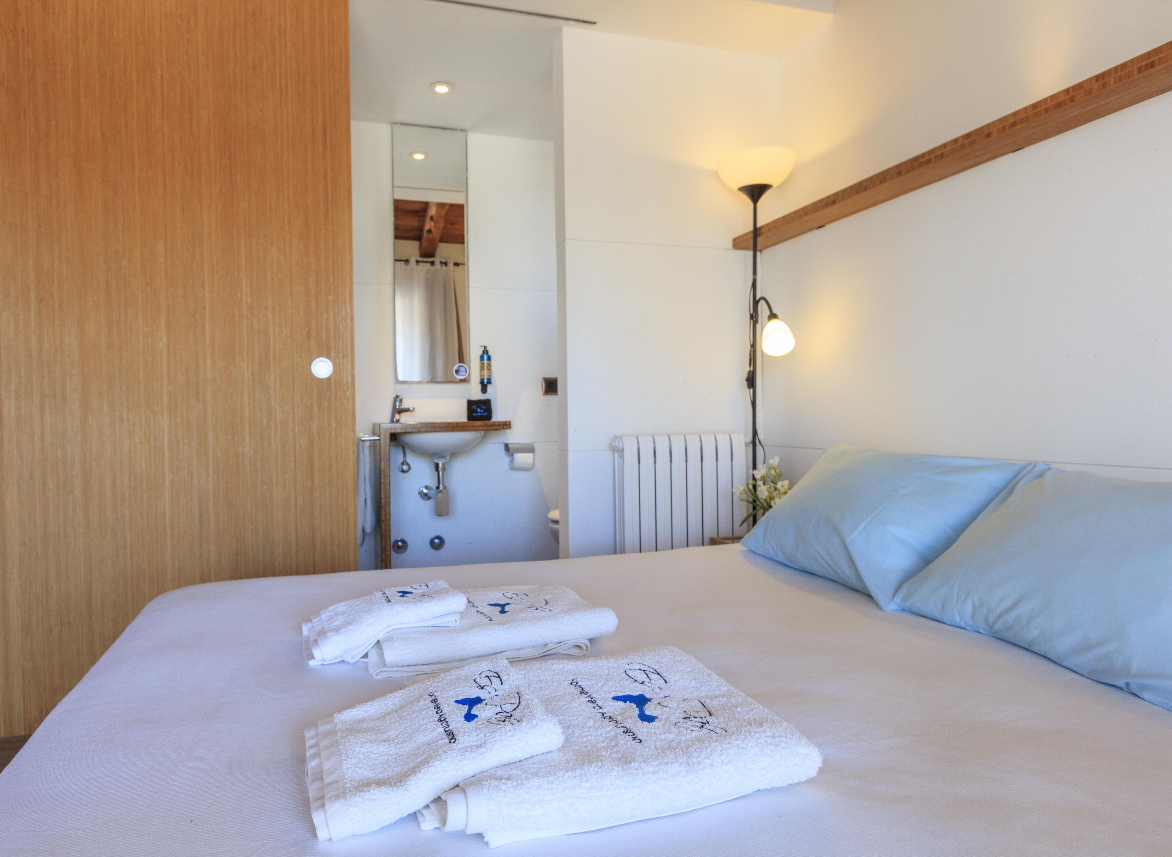 Quarto de hotel com cama, mesa de cabeceira, abajur, espelho e banheiro visível por uma porta aberta.