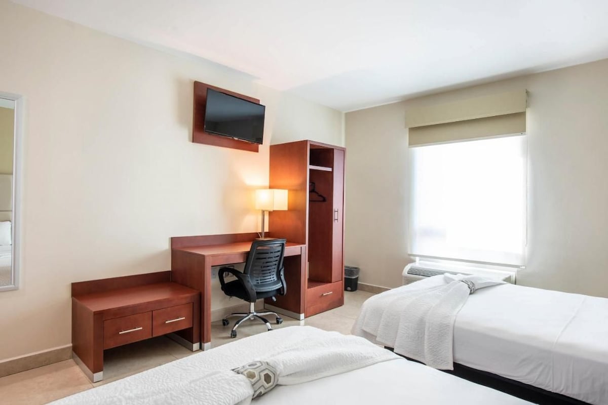 Quarto de hotel com escrivaninha, duas camas, televisão e janela.