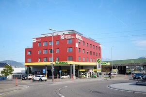 Um edifício vermelho de vários andares com uma seção inferior amarela identificada com as placas "AGROLA" e "HOTEL KRIESEL".