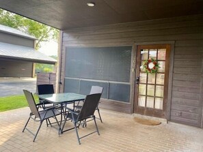 Um pátio externo com mesa e cadeiras, área coberta com uma porta e piso de tijolos.