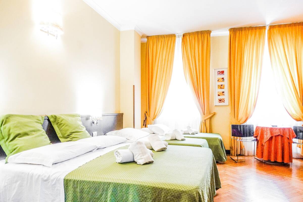 Quarto de hotel com duas camas, colchas verdes, almofadas brancas e piso de madeira.
