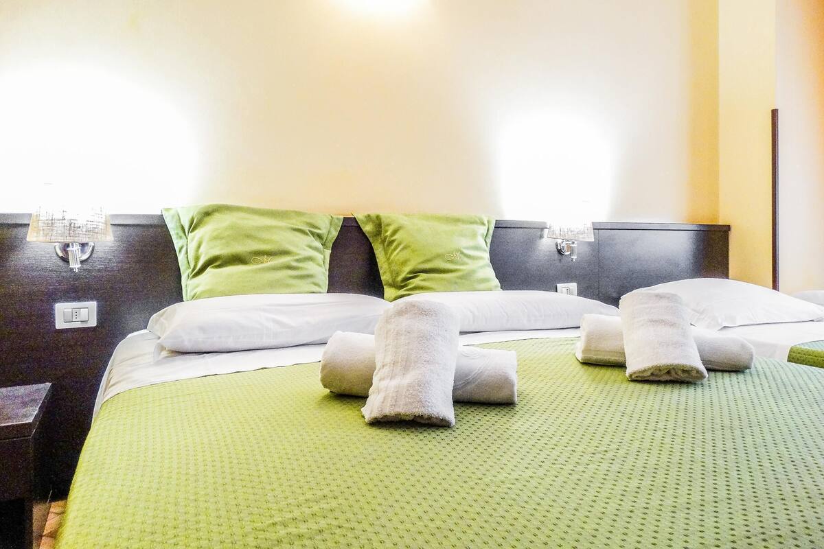 Cama bem arrumada com roupa de cama verde, duas almofadas e toalhas enroladas. Há duas mesas de cabeceira com abajures e uma luminária na parede acima da cama.