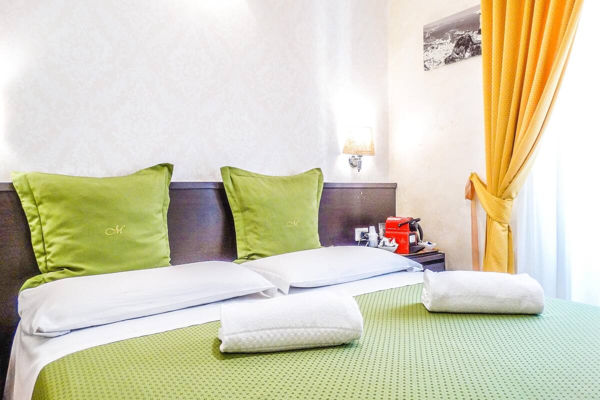 Quarto de hotel com cama, almofadas verdes, manta branca e cobre-leito verde.
