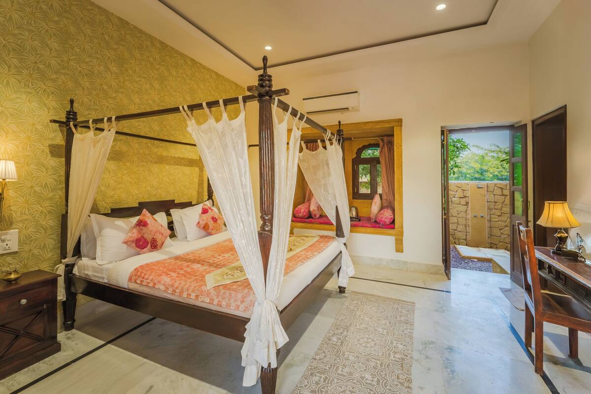 Quarto com cama de dossel, mesa de cabeceira, escrivaninha com abajur e vista para uma parede de pedra e vegetação lá fora.