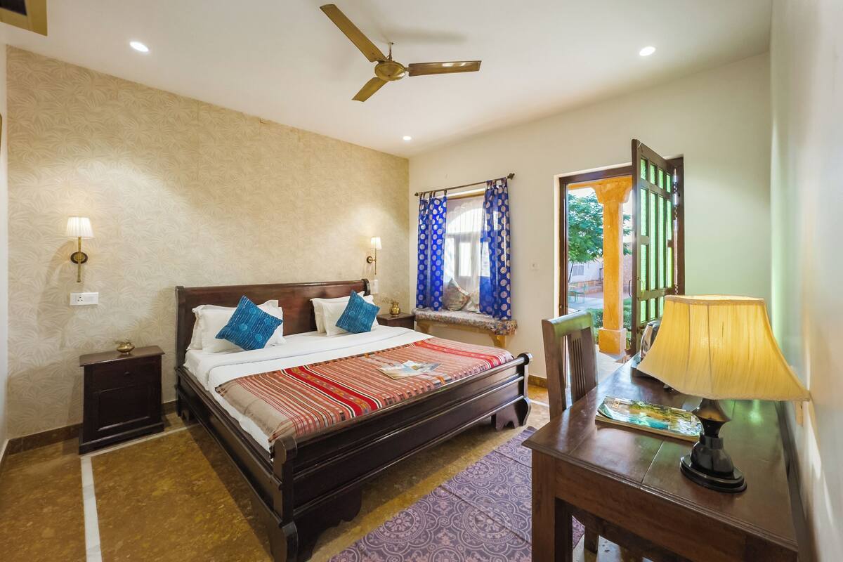 Quarto de hotel com cama, escrivaninha, cadeira, abajur, ventilador de teto e janela com cortinas.