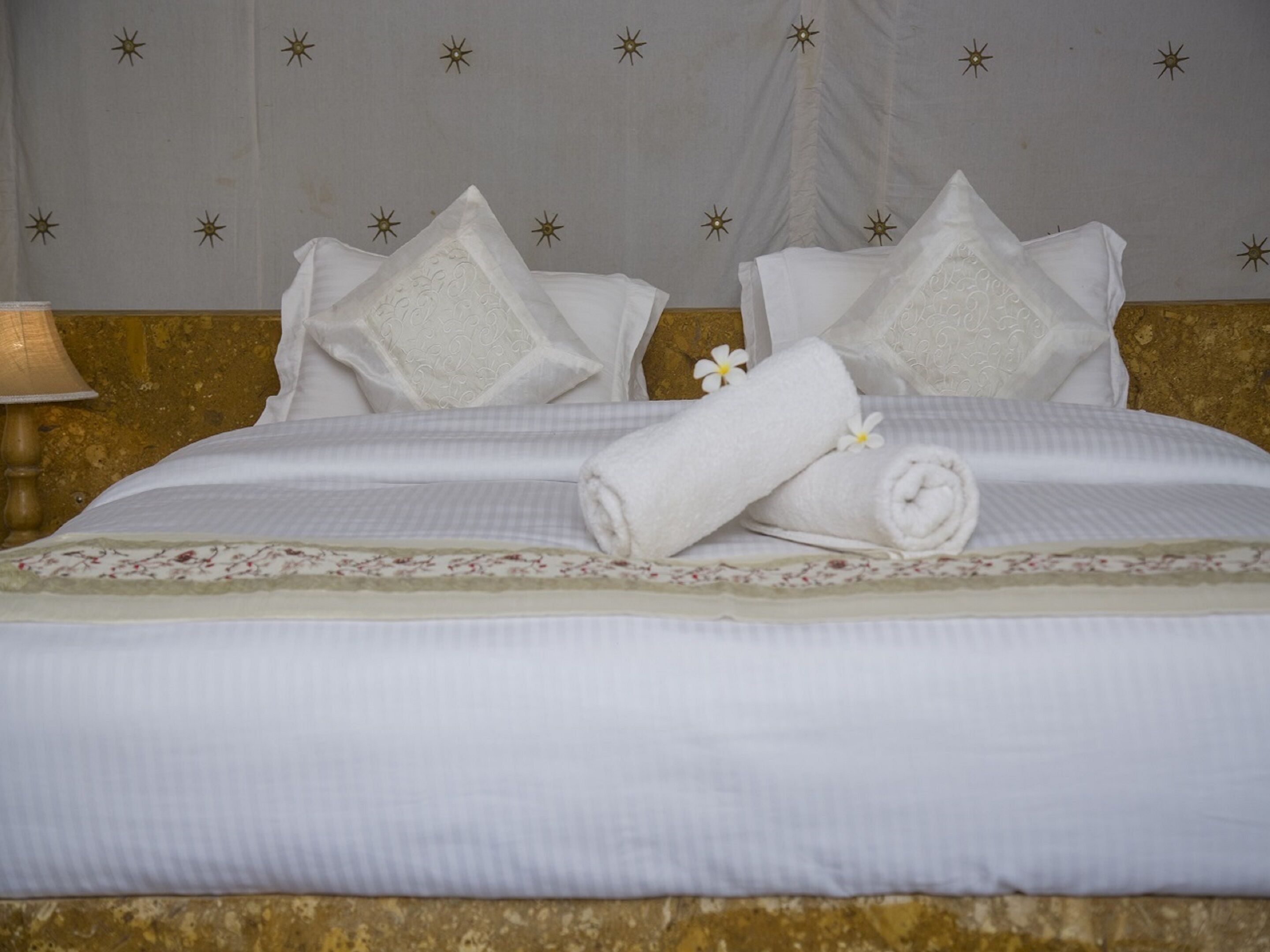 Uma cama bem arrumada com lençóis e almofadas brancos, uma toalha enrolada com uma flor e uma luminária na lateral.