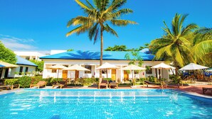 Piscina com espreguiçadeiras e guarda-sóis, cercada por palmeiras e um edifício com telhado azul.