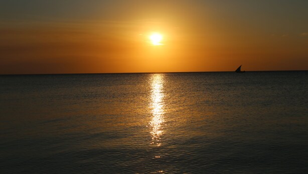 Um pôr do sol sobre um mar calmo, com um veleiro ao longe.