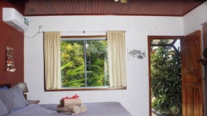 Quarto com cama, janela com cortinas, ar condicionado fixado na parede e porta que dá para uma varanda com plantas.